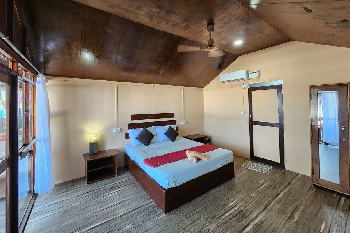 Quarto com teto de madeira, cama com roupa de cama azul e branca, mesa de cabeceira, ar condicionado e uma porta.