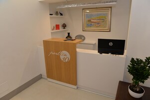 Um balcão de recepção em madeira, um quadro emoldurado acima e um vaso com planta à direita.