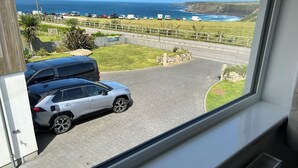 Vista da janela para uma área de estacionamento com um SUV branco e uma van camper, uma área gramada e uma paisagem costeira com falésias e o mar.