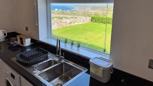 Uma cozinha com uma pia de aço inoxidável, uma chaleira e uma cafeteira. Há uma janela com vista para um gramado e um muro de pedra.