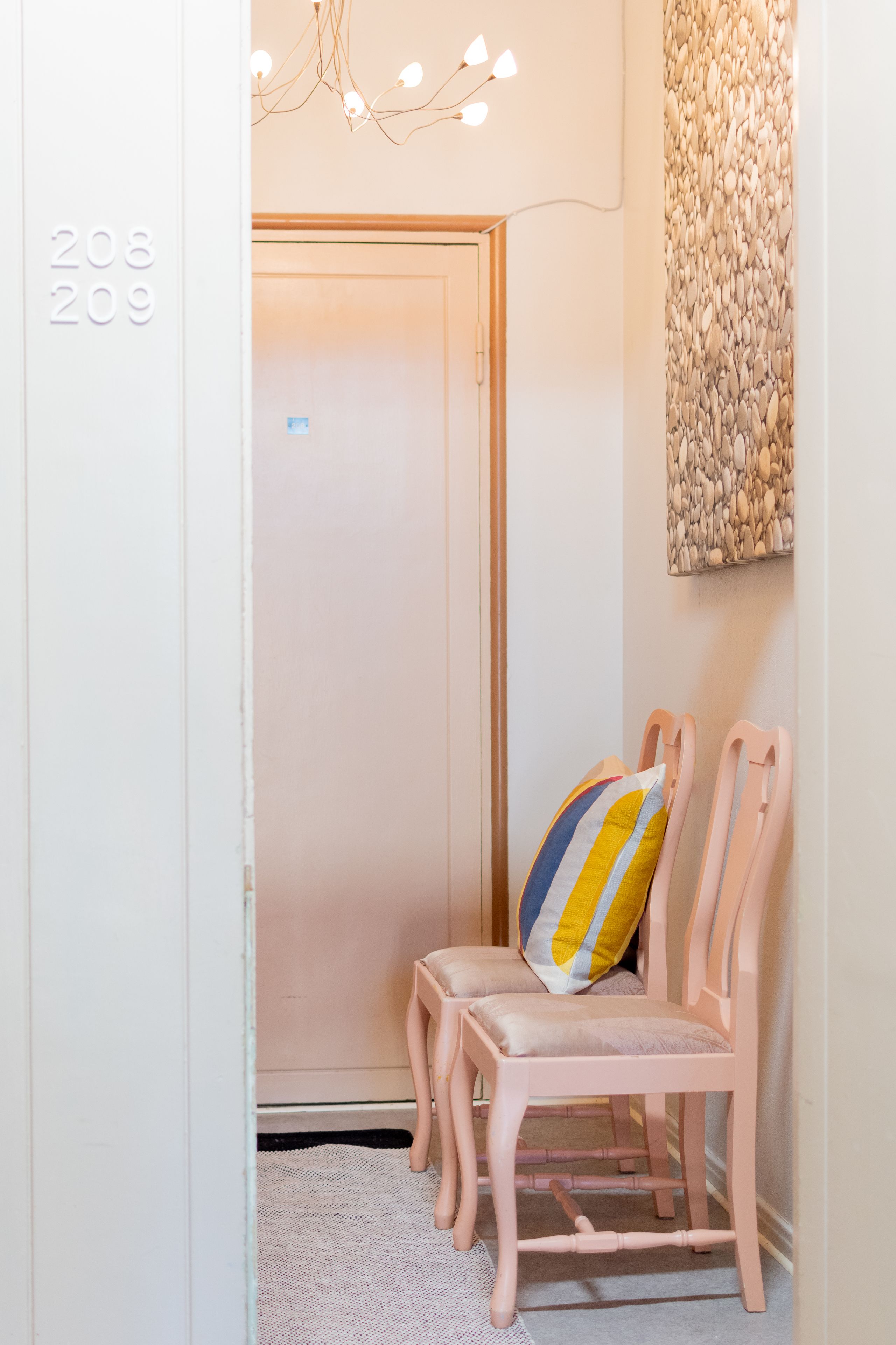 Quarto com porta branca numerada, cadeira rosa com almofada listrada e obra de arte com textura na parede.