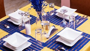 Mesa de jantar posta com pratos brancos, talheres e copos de vidro transparente, incluindo copos de vinho e copos de água, sobre toalha de mesa amarela com listras azuis.