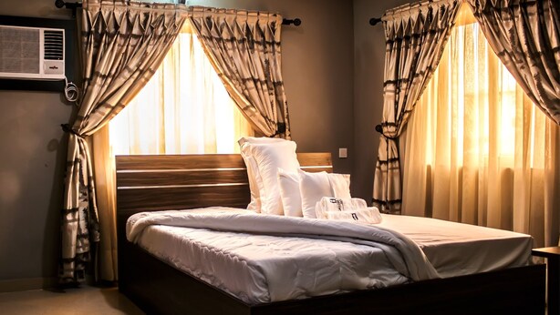Cama bem arrumada com lençóis e almofadas brancas, cabeceira de madeira e mesa de cabeceira com luminária.