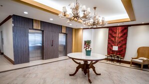 Um hall de entrada com uma mesa redonda, dois elevadores, uma tapeçaria vermelha e uma cadeira de madeira.