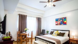 Quarto de hotel com cama, escrivaninha com cadeira, televisão e ventoinha no teto.