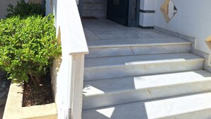 Entrada de um edifício com escadaria que leva a uma porta, uma pequena área ajardinada e um corrimão visível.