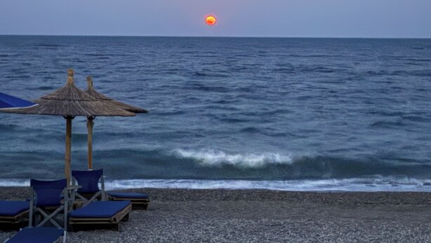 Uma praia com uma fileira de cadeiras azuis e um guarda-sol de palha.
