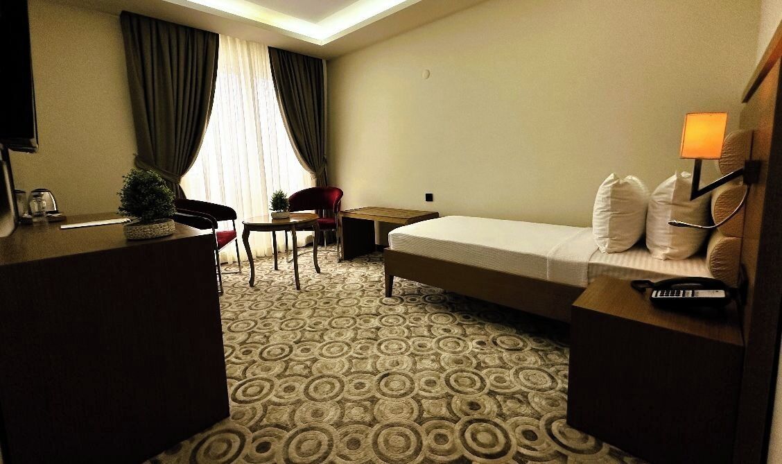Quarto de hotel com uma cama, uma secretária com cadeira, uma pequena mesa com uma planta, um candeeiro e um comando de televisão num criado-mudo.