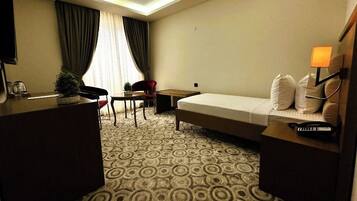 Quarto de hotel com uma cama, uma secretária com cadeira, uma pequena mesa com uma planta, um candeeiro e um comando de televisão num criado-mudo.