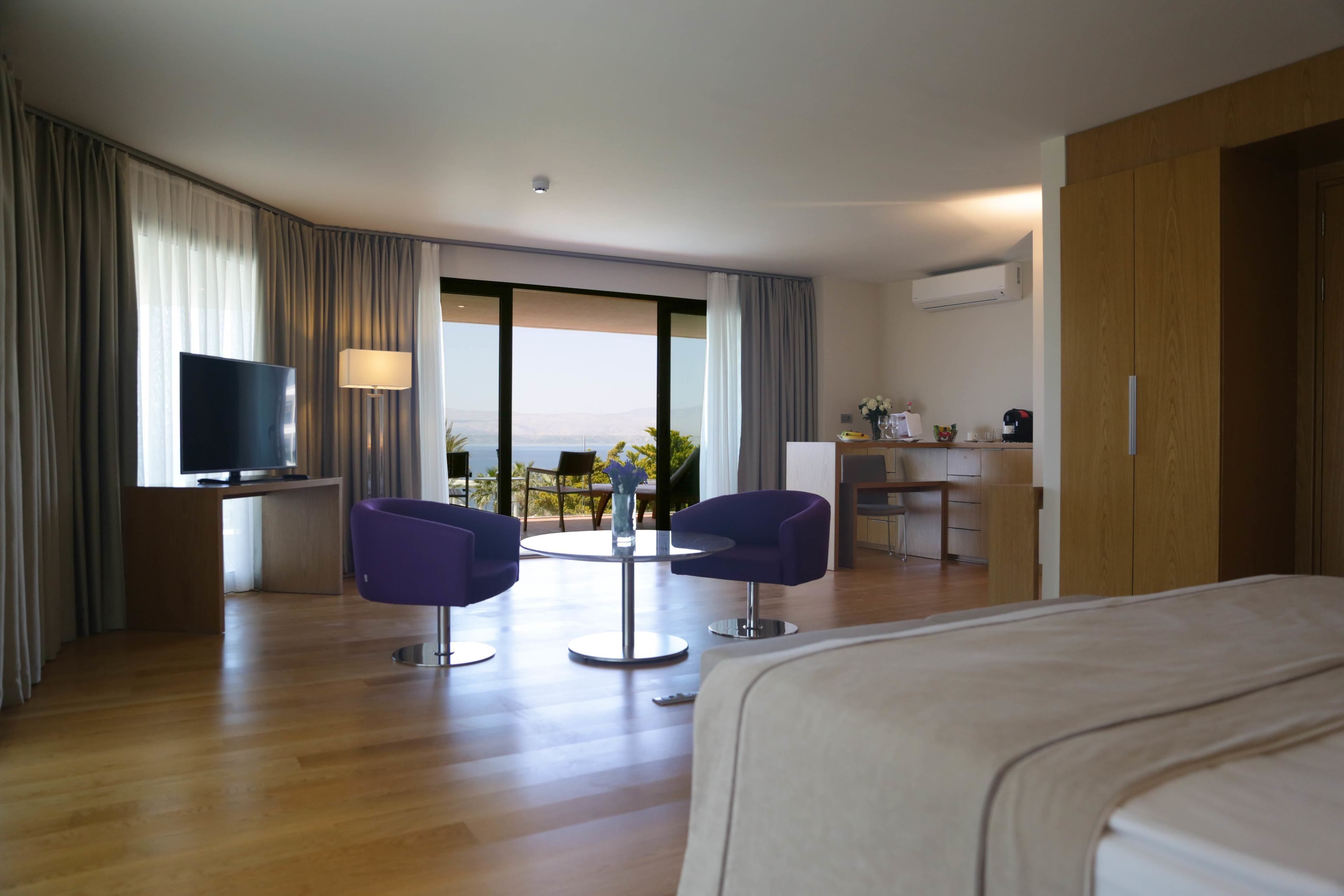 Quarto de hotel moderno com uma cama grande, televisão, duas cadeiras roxas, mesa de jantar e vista para o exterior.