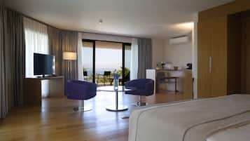 Quarto de hotel moderno com uma cama grande, televisão, duas cadeiras roxas, mesa de jantar e vista para o exterior.