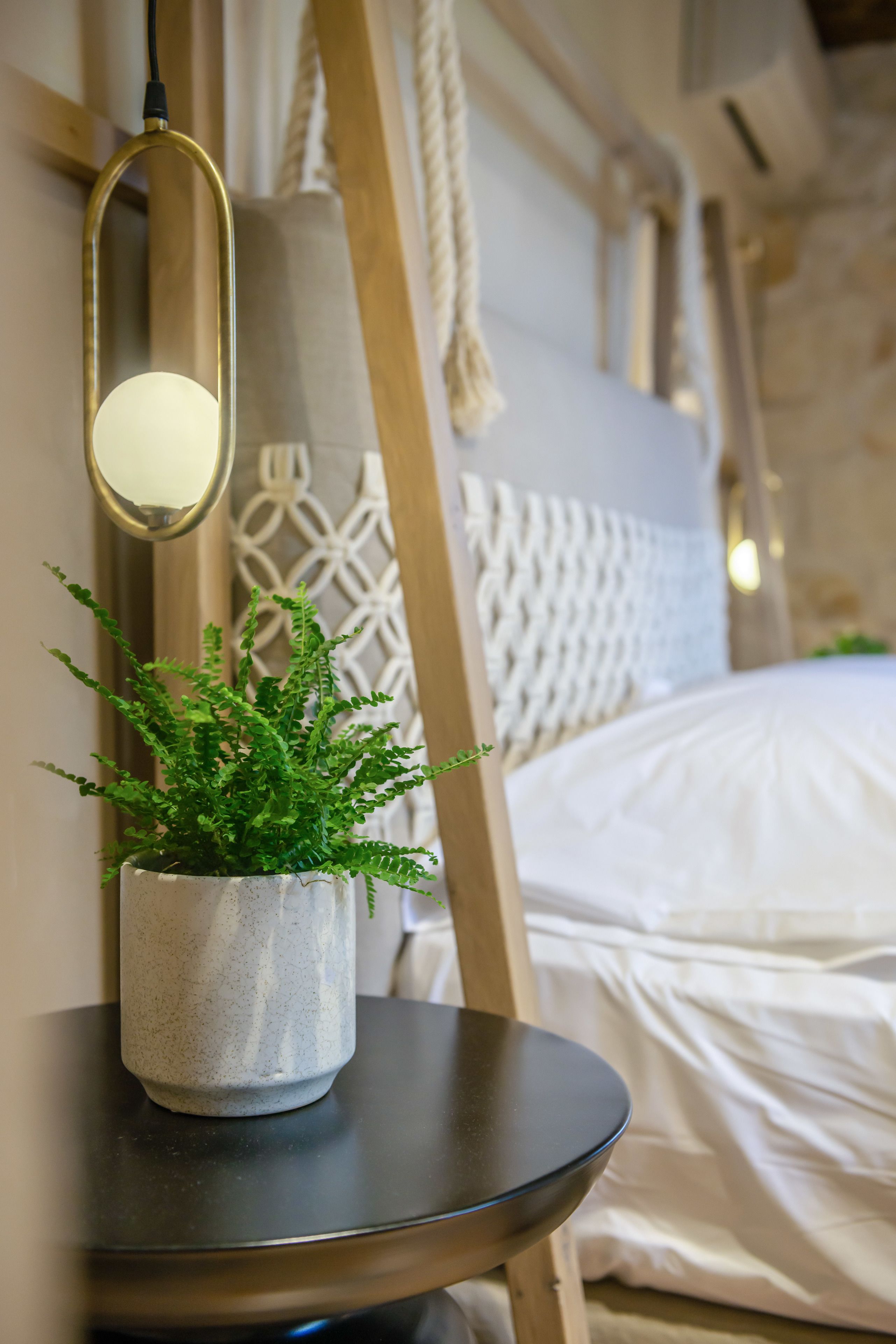 Uma cama com roupa de cama branca, uma mesa redonda com um vaso de planta e um espelho pendurado.