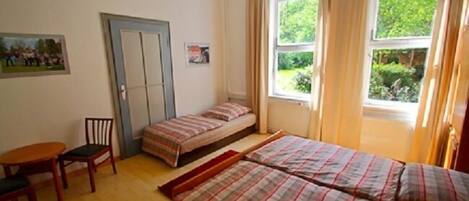 Quarto com duas camas, uma mesinha e uma cadeira. Há uma janela com cortinas, uma porta e um quadro na parede.