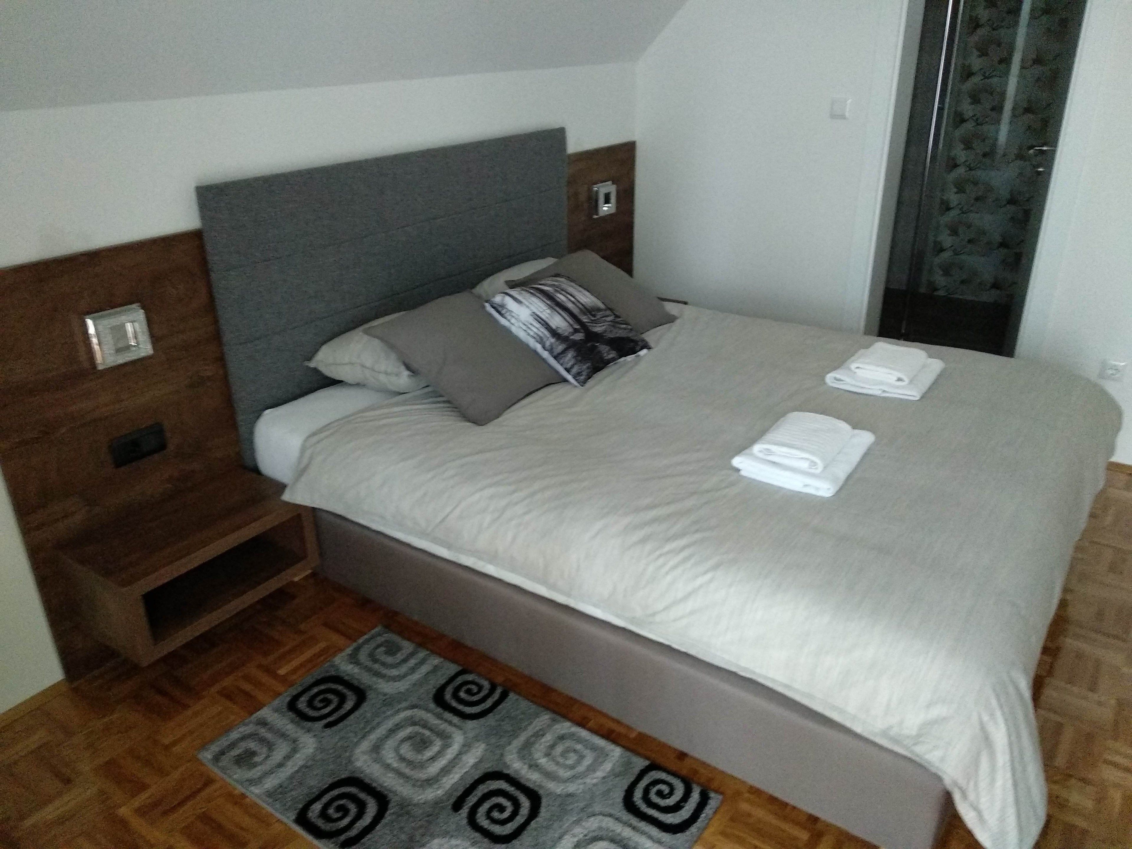 Uma cama bem arrumada com cabeceira cinza, tapete cinza e preto com padrão, e duas toalhas brancas na cama.