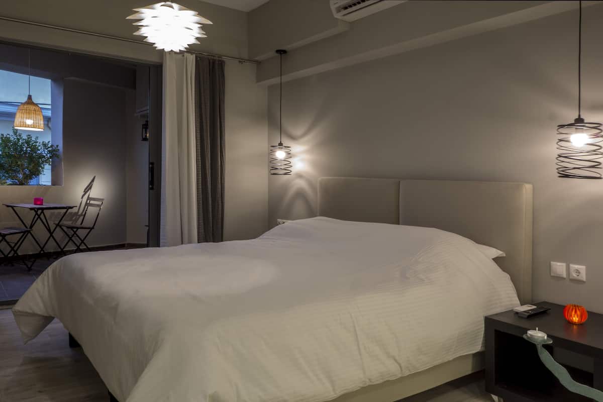 Quarto moderno com uma cama grande, uma mesinha de cabeceira com abajur e vista para uma varanda com mesa e cadeiras.