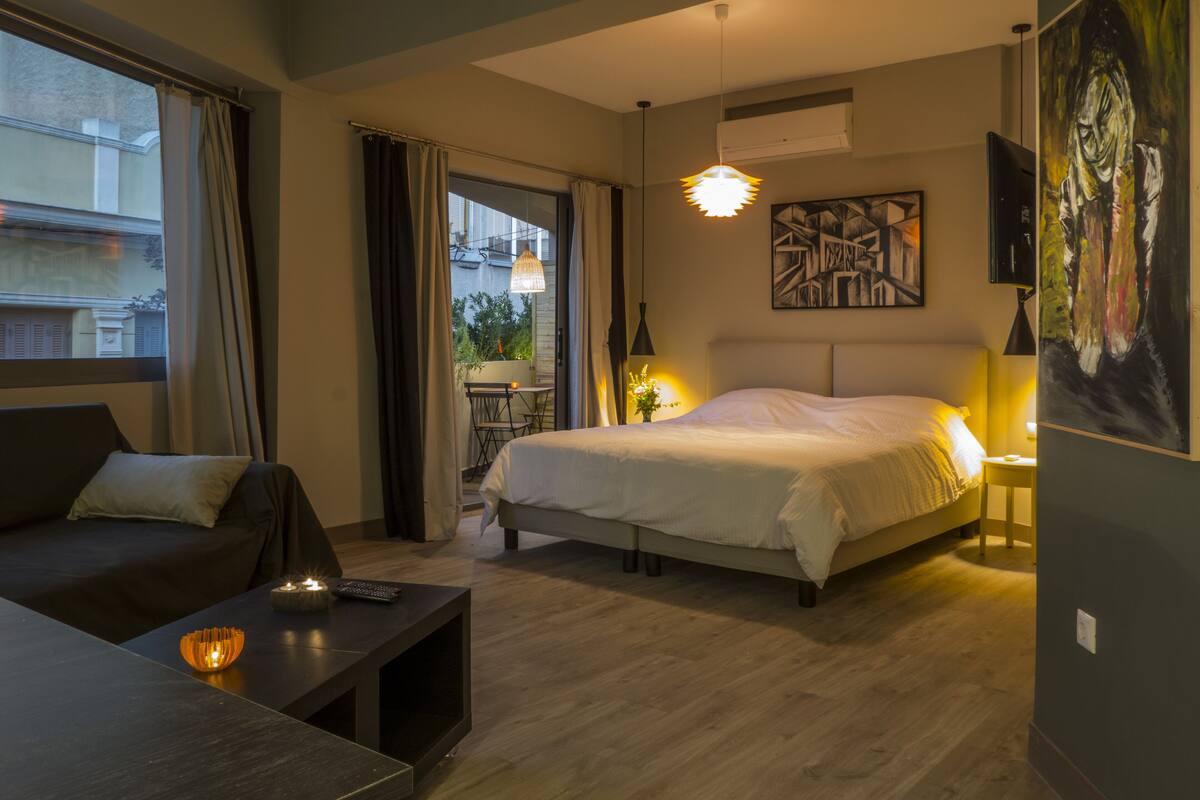 Quarto moderno com cama, sofá, mesa de cabeceira com luminária e vista para o exterior.
