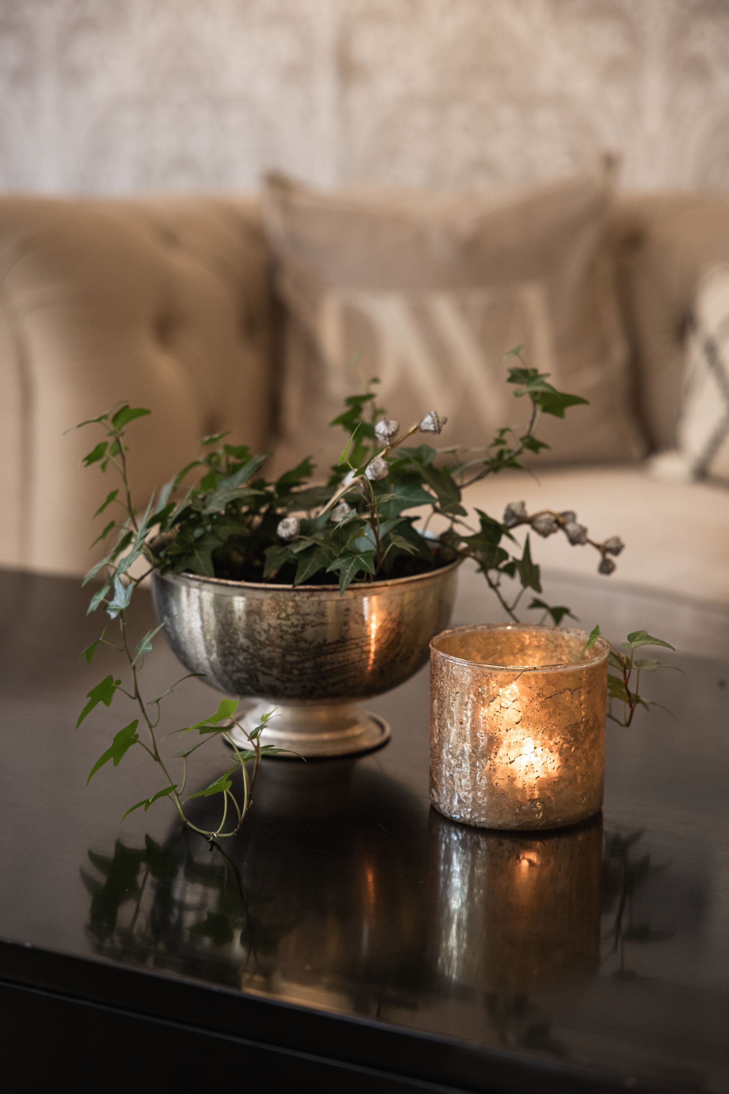 Uma tigela metálica com plantas verdes sobre uma superfície refletora, uma vela e um sofá bege ao fundo.