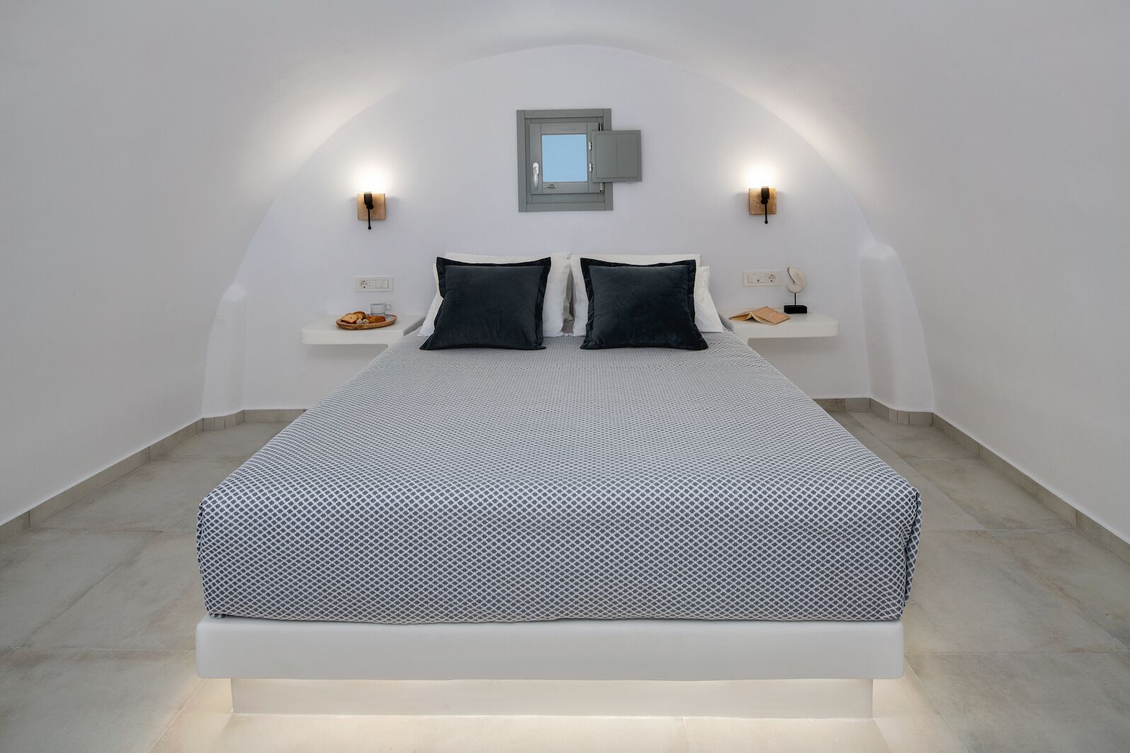 Um quarto moderno com uma cama, duas mesas de cabeceira, um criado-mudo, uma luminária na parede e uma pequena janela.