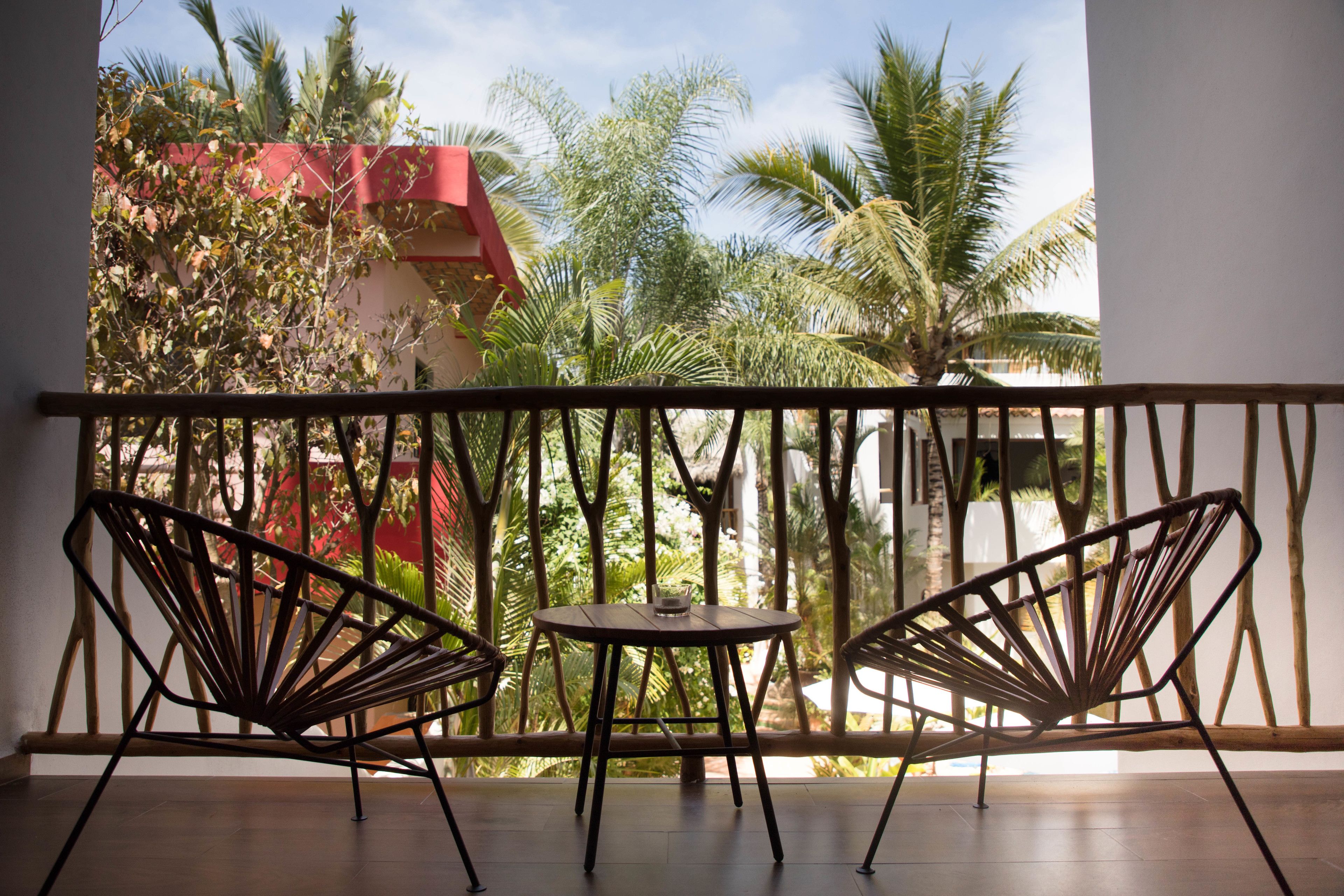 Uma varanda com duas cadeiras e uma pequena mesa, com vista para um jardim com palmeiras e um prédio com telhado vermelho.