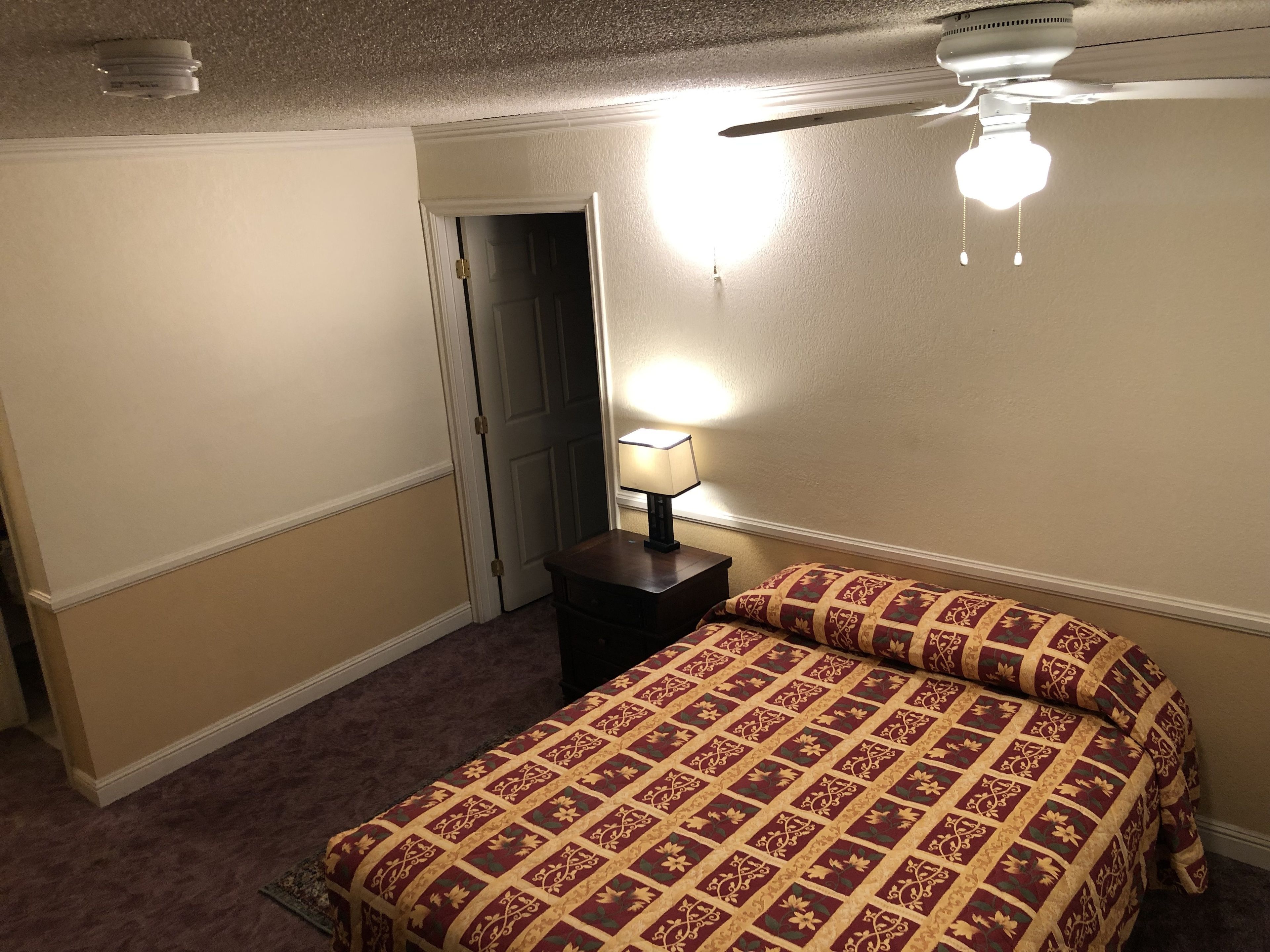 Quarto com cama, mesa de cabeceira, luminária, ventilador de teto e porta.