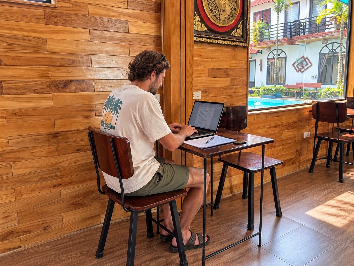 Uma pessoa sentada à mesa de madeira com um laptop, trabalhando em um quarto com paredes de madeira e uma janela com vista para uma piscina.