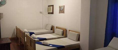 Quarto de hotel com três camas de solteiro, um banco de madeira, uma mesa pequena e uma cortina azul.