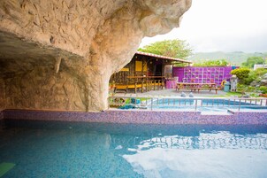 Uma área de piscina com uma formação rochosa, um edifício roxo com telhado vermelho e vista para o verde ao fundo.