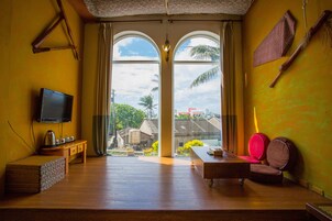 Quarto com televisão, mobiliário de madeira, um banco e vista para prédios e palmeiras através de grandes janelas.