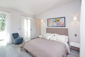 Quarto moderno com uma cama grande, uma cadeira, uma mesinha de cabeceira e uma porta que dá para uma varanda.