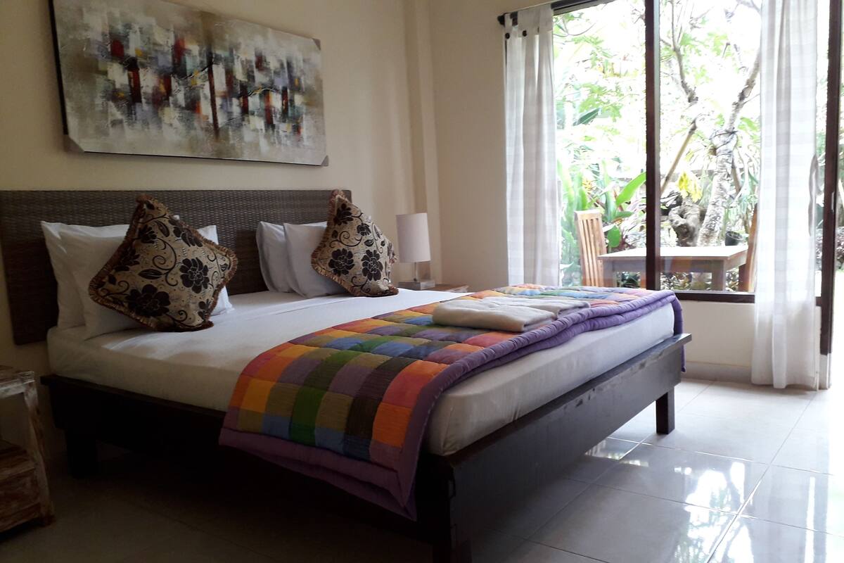 Uma cama bem arrumada com um edredom colorido, cabeceira com almofadas decorativas, um quadro na parede e vista para o verde pela janela.