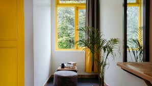 Um quarto com porta amarela, janela com vista para vegetação, um banquinho, mesa com livros e caneca, e um vaso de planta.
