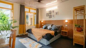 Quarto com cama, mesas de cabeceira, cabeceira de madeira, janela com cortinas, um vaso de planta e vista para o verde lá fora.