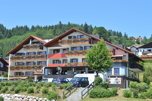 Hotel de vários andares com varandas, área de estacionamento e montanhas ao fundo.