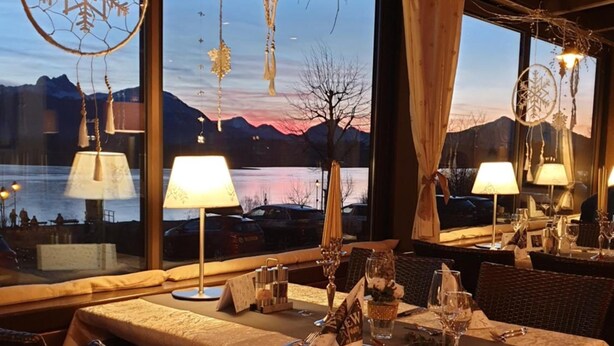 Restaurante com vista para montanhas e um lago, com mesas preparadas, incluindo copos de vinho e um arranjo de velas no centro.