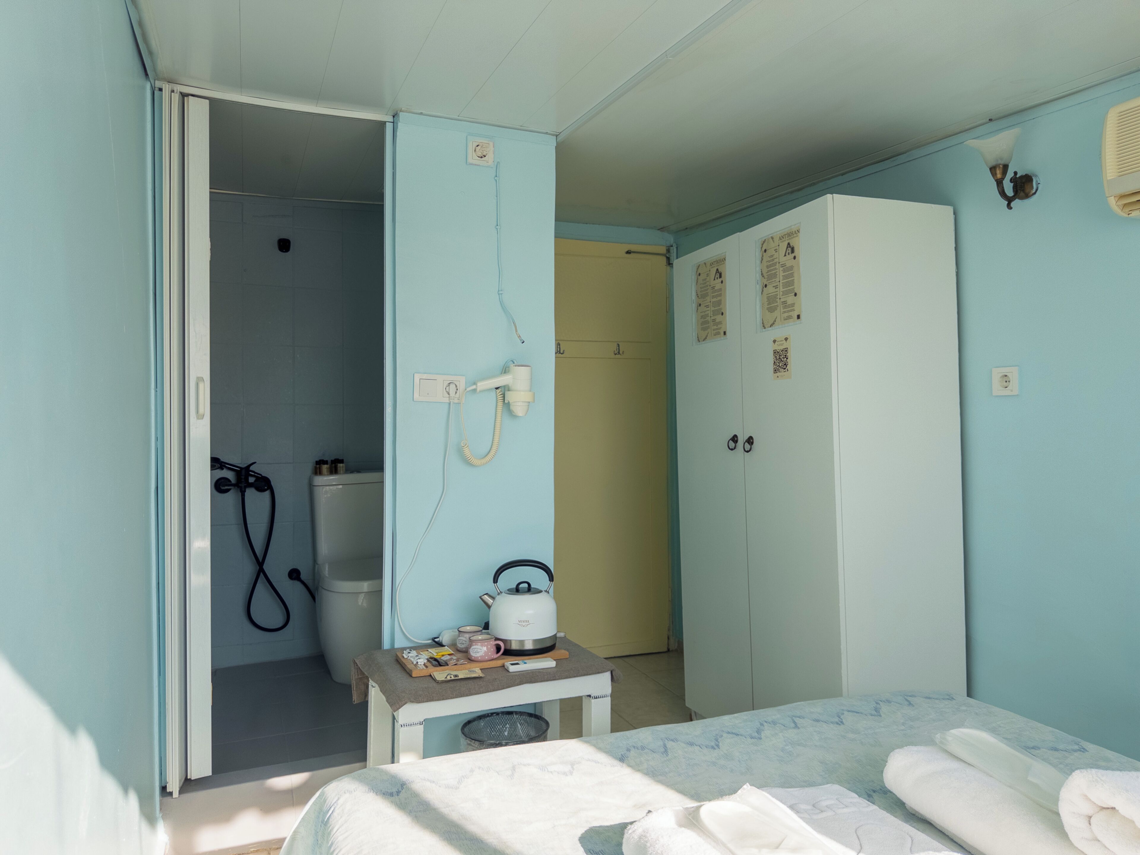 Quarto com cama, uma mesinha com chaleira, guarda-roupa e banheiro com chuveiro e vaso sanitário.