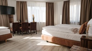 Quarto de hotel moderno com uma cama grande, uma mesa com cadeiras, televisão de tela plana e janelas amplas com cortinas.