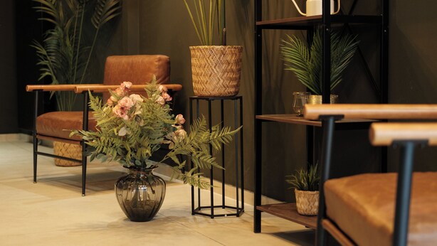 Uma sala de estar moderna com um sofá de couro marrom, um vaso de vidro com flores, uma cesta de vime e uma pequena planta em vaso sobre um suporte de metal preto.
