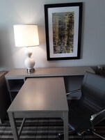 Quarto de hotel com uma mesa branca, uma cadeira preta, uma luminária e um quadro abstrato na parede.
