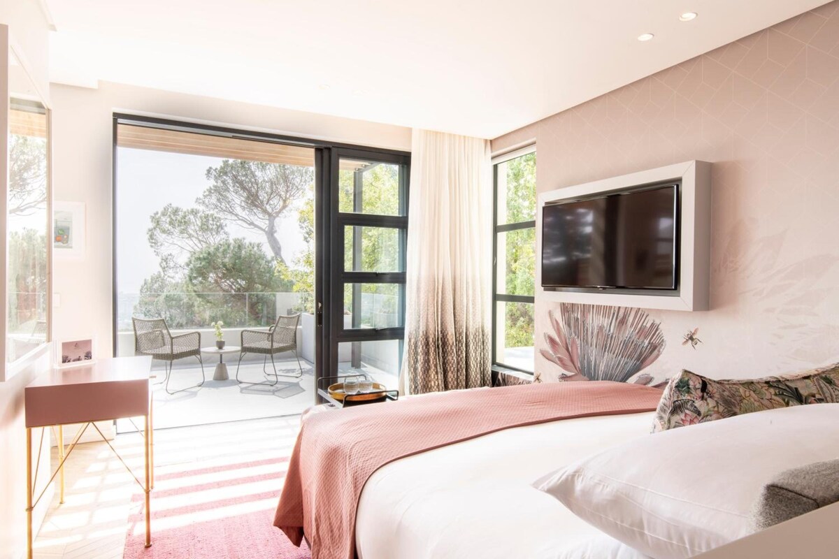 Quarto com uma cama grande, televisão na parede, varanda com móveis de exterior e porta de vidro deslizante.