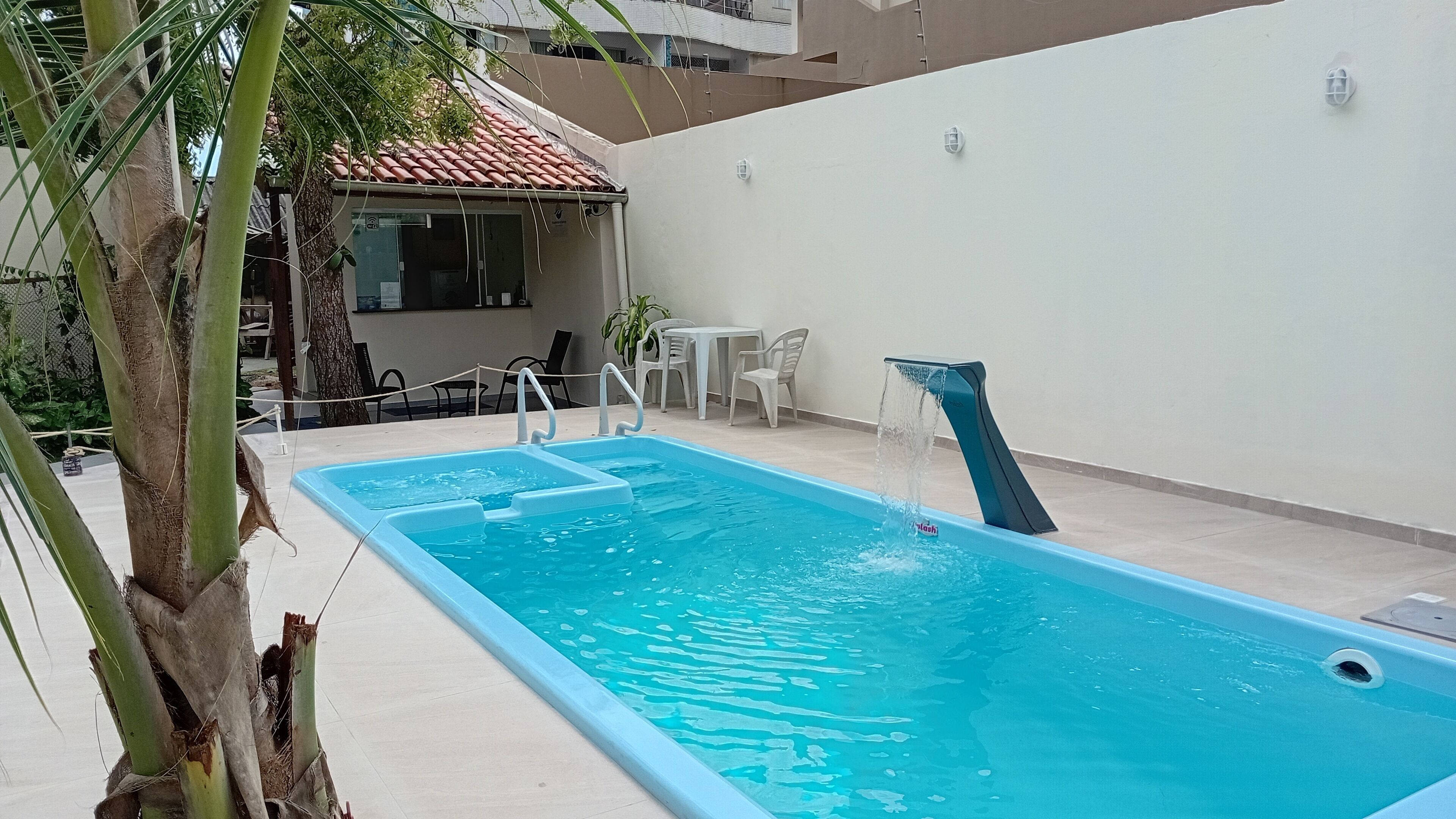 Uma piscina com trampolim, cercada por um muro branco, uma mesa pequena e cadeiras.
