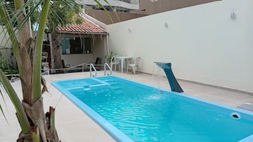 Uma piscina com trampolim, cercada por um muro branco, uma mesa pequena e cadeiras.