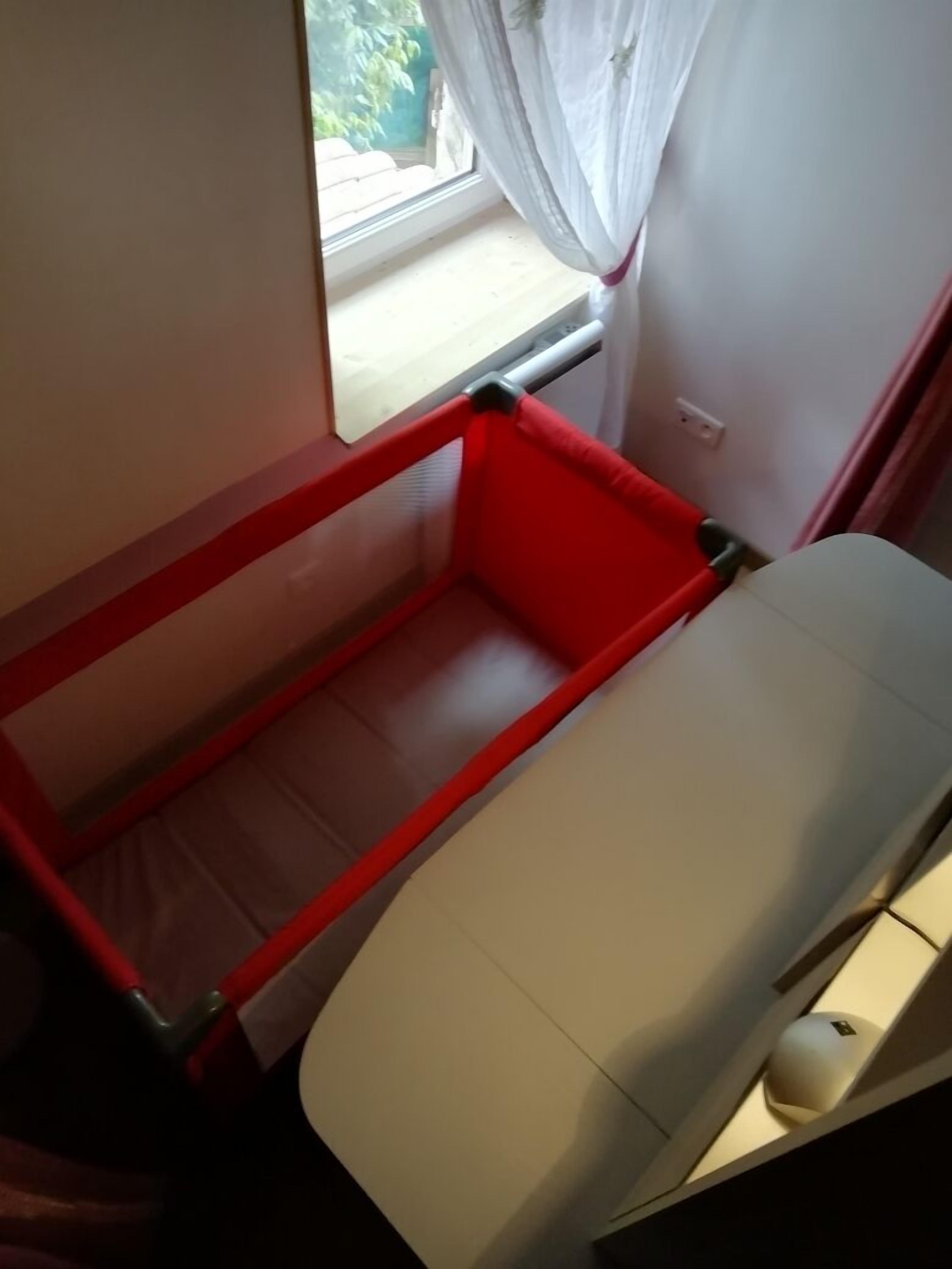 Quarto com cama vermelha e branca, janela com cortinas e vista para o verde lá fora.