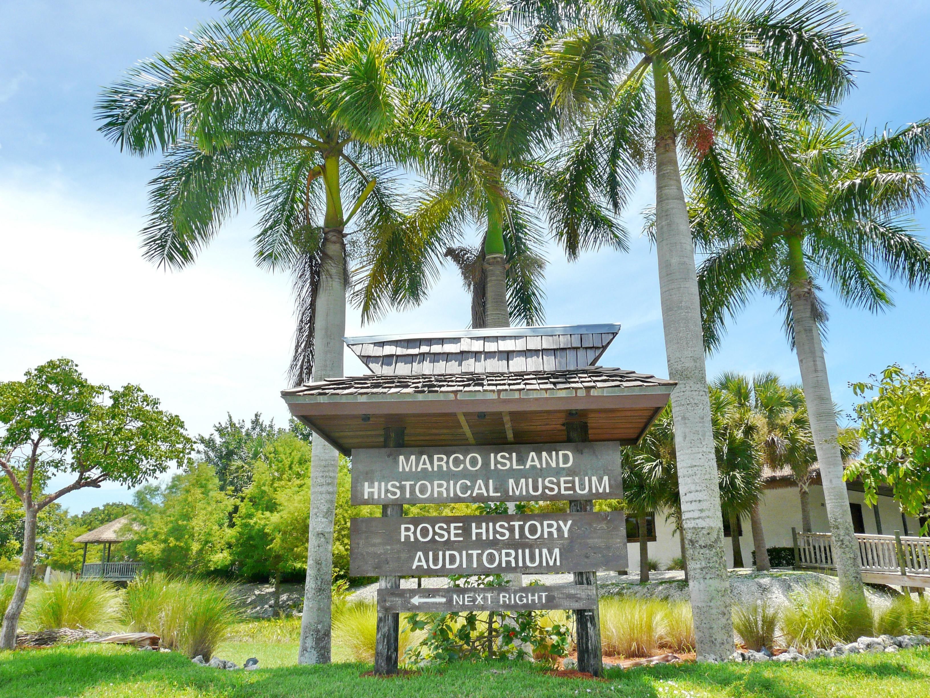 Placa do Museu Histórico de Marco Island, com uma estrutura de madeira e palmeiras ao fundo.