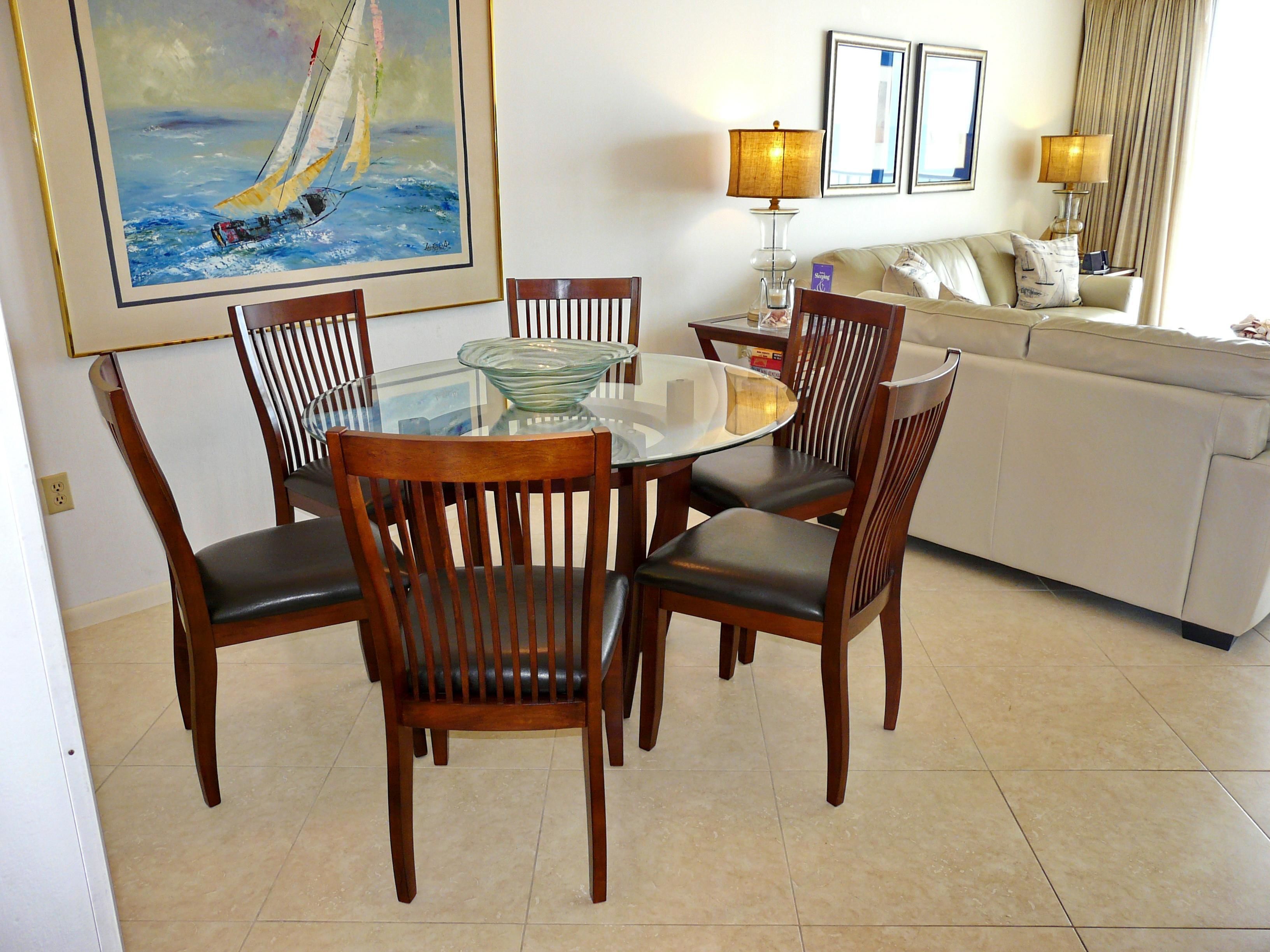 Uma área de refeições com mesa de vidro cercada por cadeiras de madeira, um sofá e uma pintura de um veleiro.