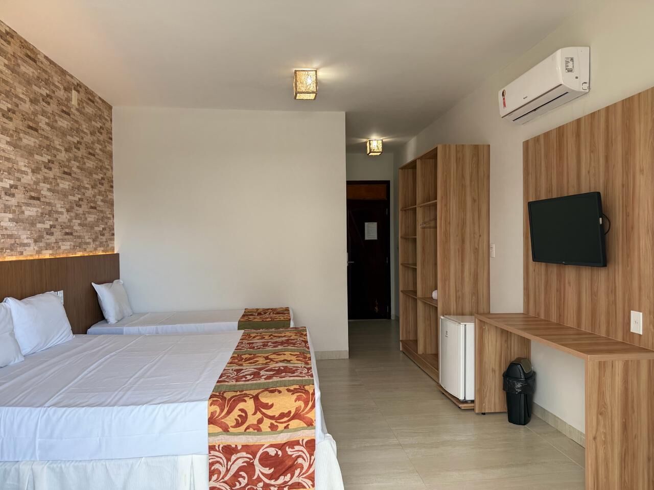 Quarto de hotel com duas camas, televisão, guarda-roupa de madeira e uma pequena cozinha.