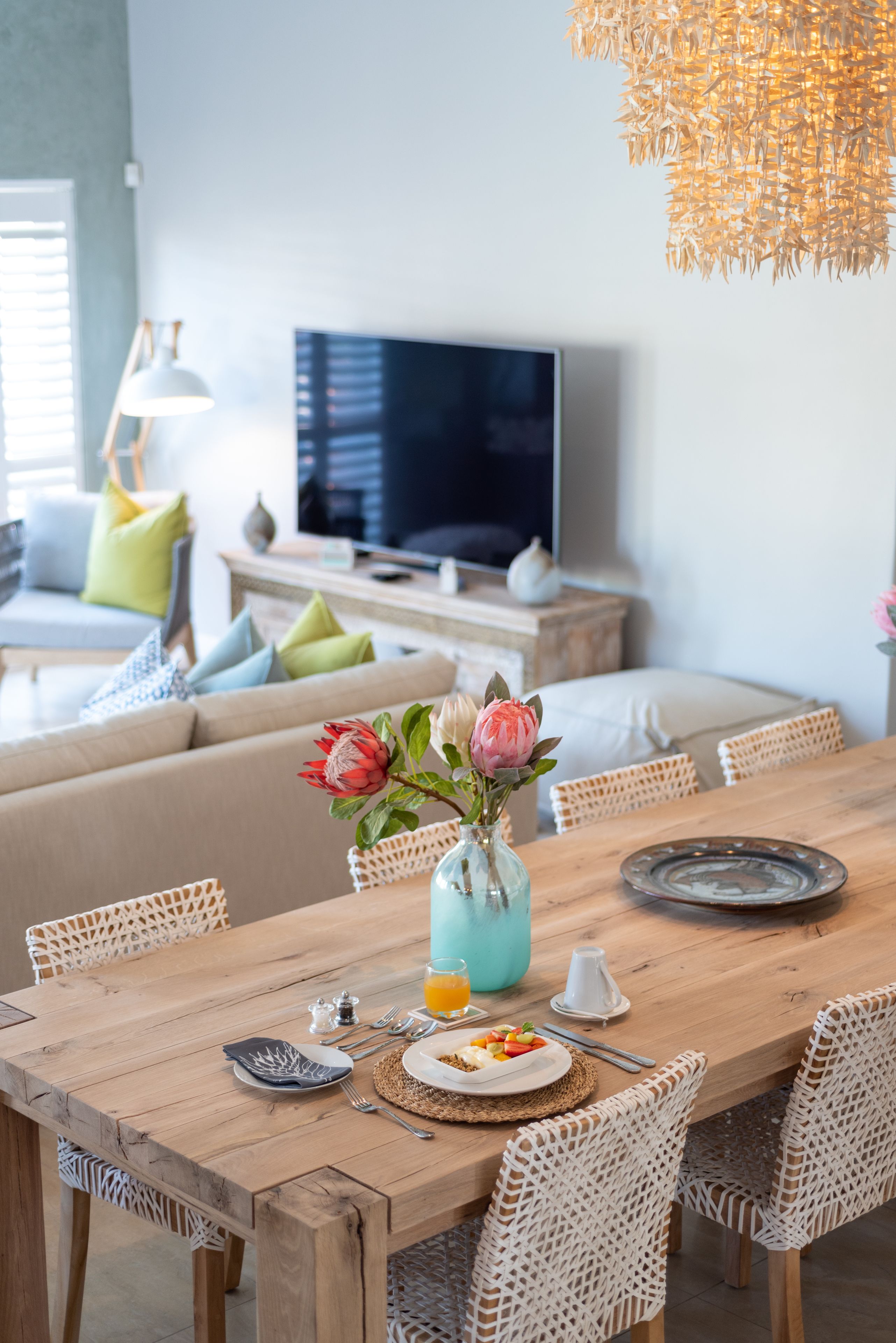 Uma sala de jantar com uma mesa de madeira posta para quatro pessoas, rodeada por cadeiras de vime branco. Uma televisão está montada na parede ao fundo, e um sofá com almofadas é visível na sala de estar.