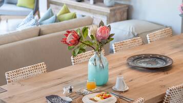 Uma sala de jantar com uma mesa de madeira posta para quatro pessoas, rodeada por cadeiras de vime branco. Uma televisão está montada na parede ao fundo, e um sofá com almofadas é visível na sala de estar.