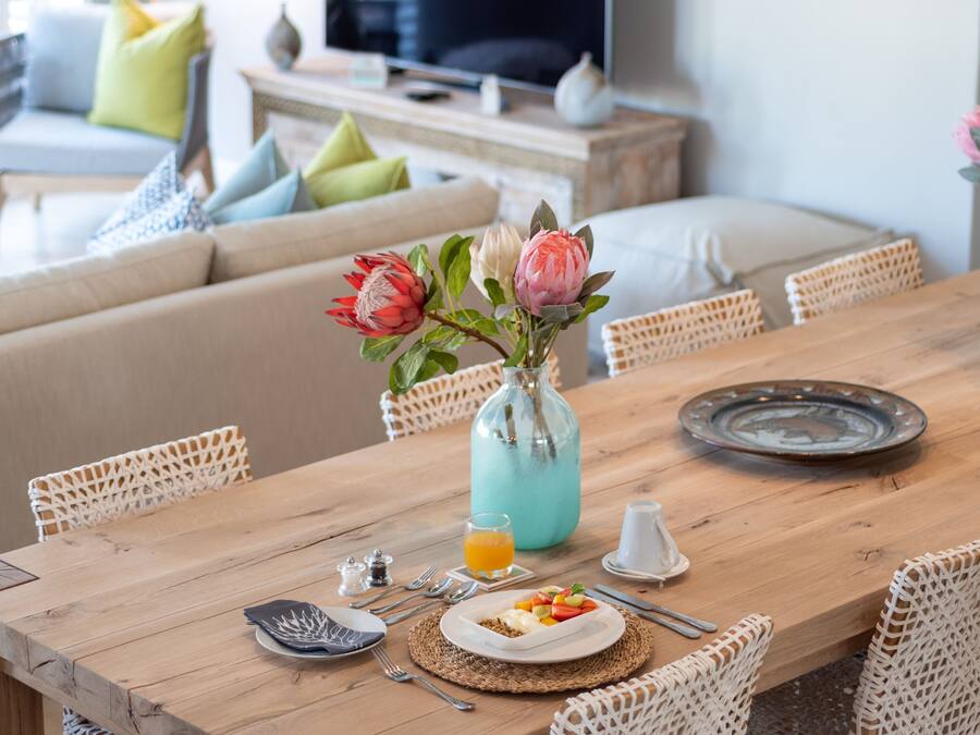 Uma sala de jantar com uma mesa de madeira posta para quatro pessoas, rodeada por cadeiras de vime branco. Uma televisão está montada na parede ao fundo, e um sofá com almofadas é visível na sala de estar.