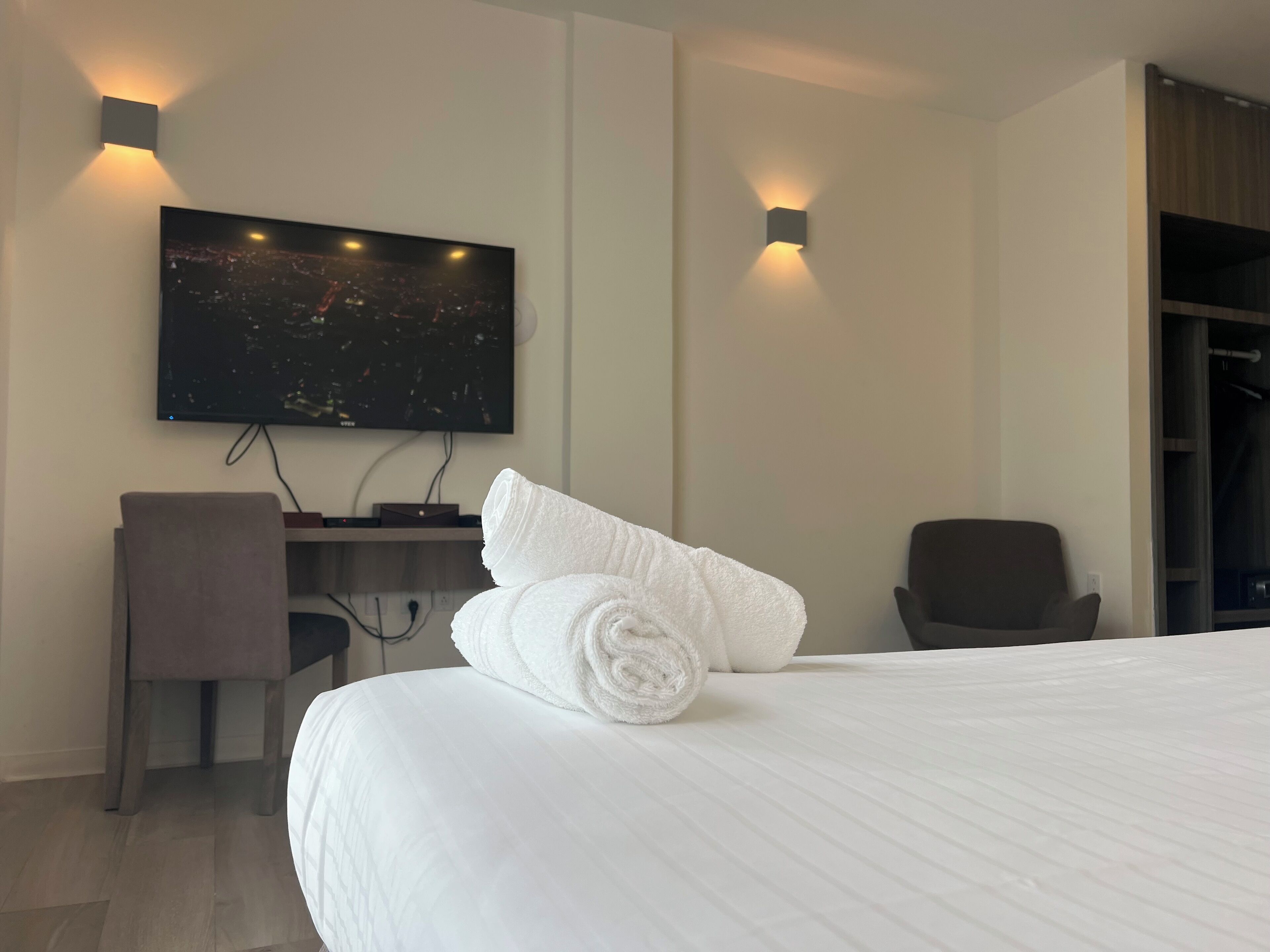 Quarto de hotel com uma cama, escrivaninha, cadeira, televisão fixada na parede e um guarda-roupa.