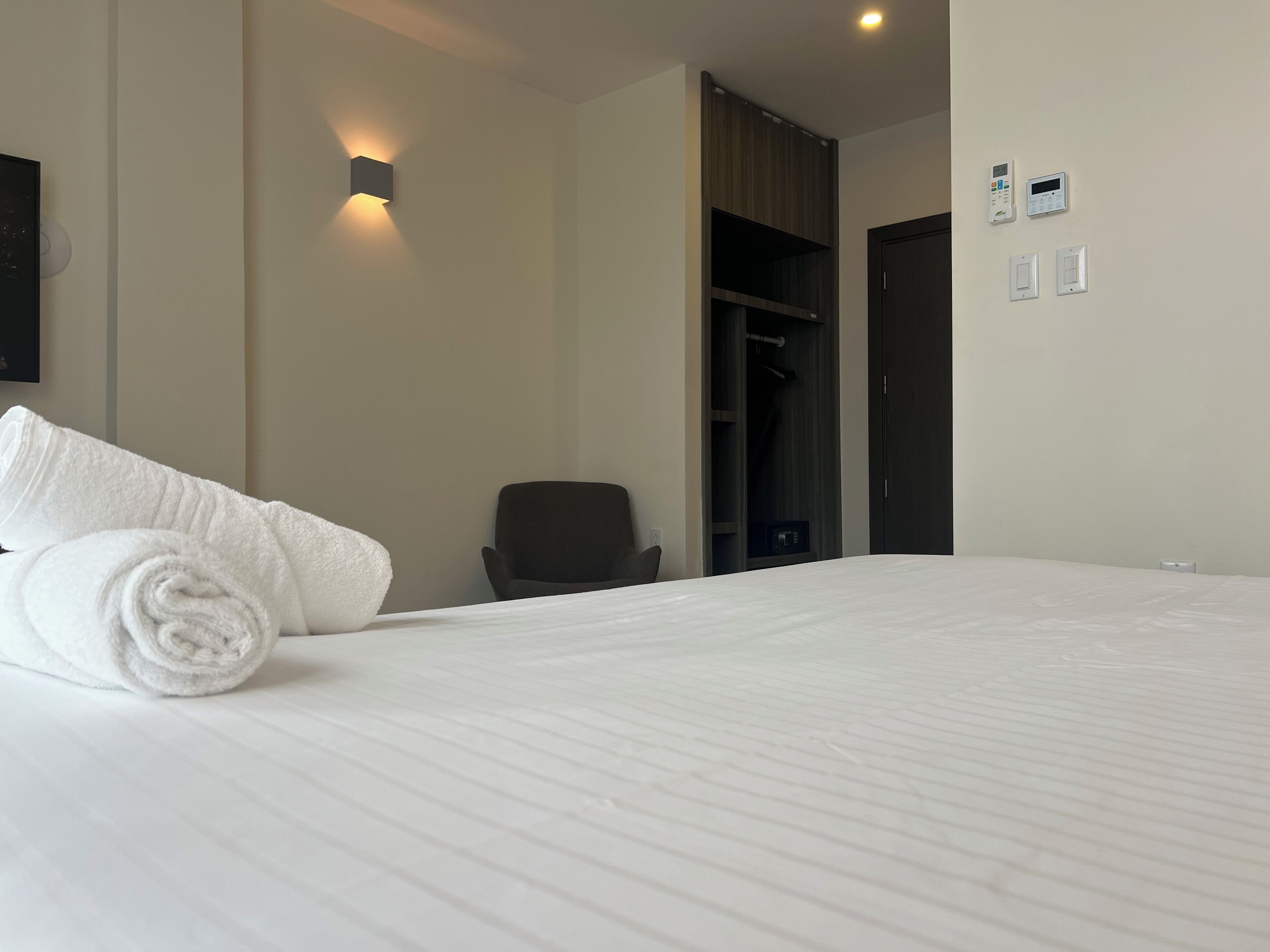 Quarto de hotel com cama bem arrumada, uma cadeira, guarda-roupa e termostato fixado na parede.